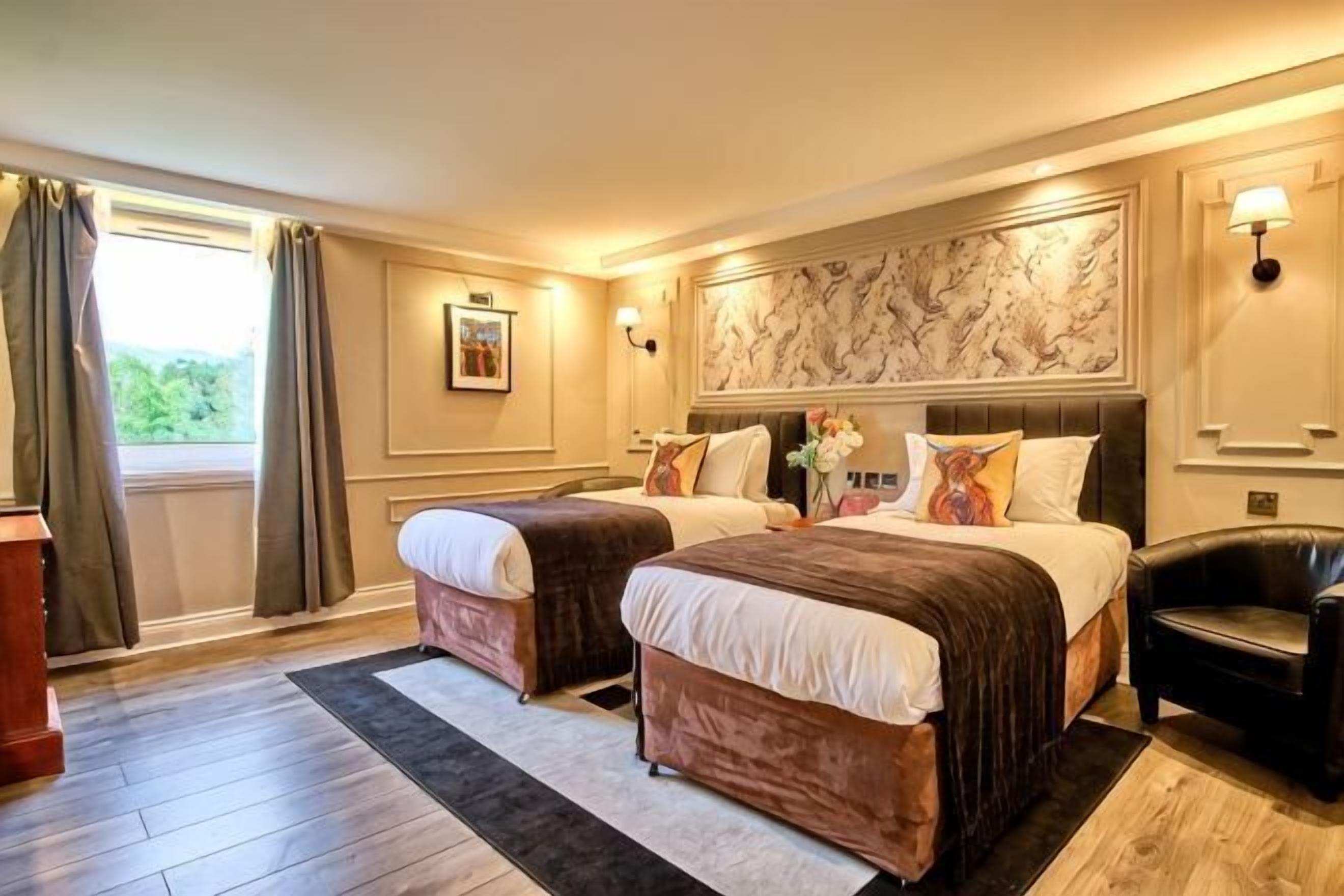 Quarto de hotel com duas camas, uma janela grande, parede de mármore e uma poltrona de couro.