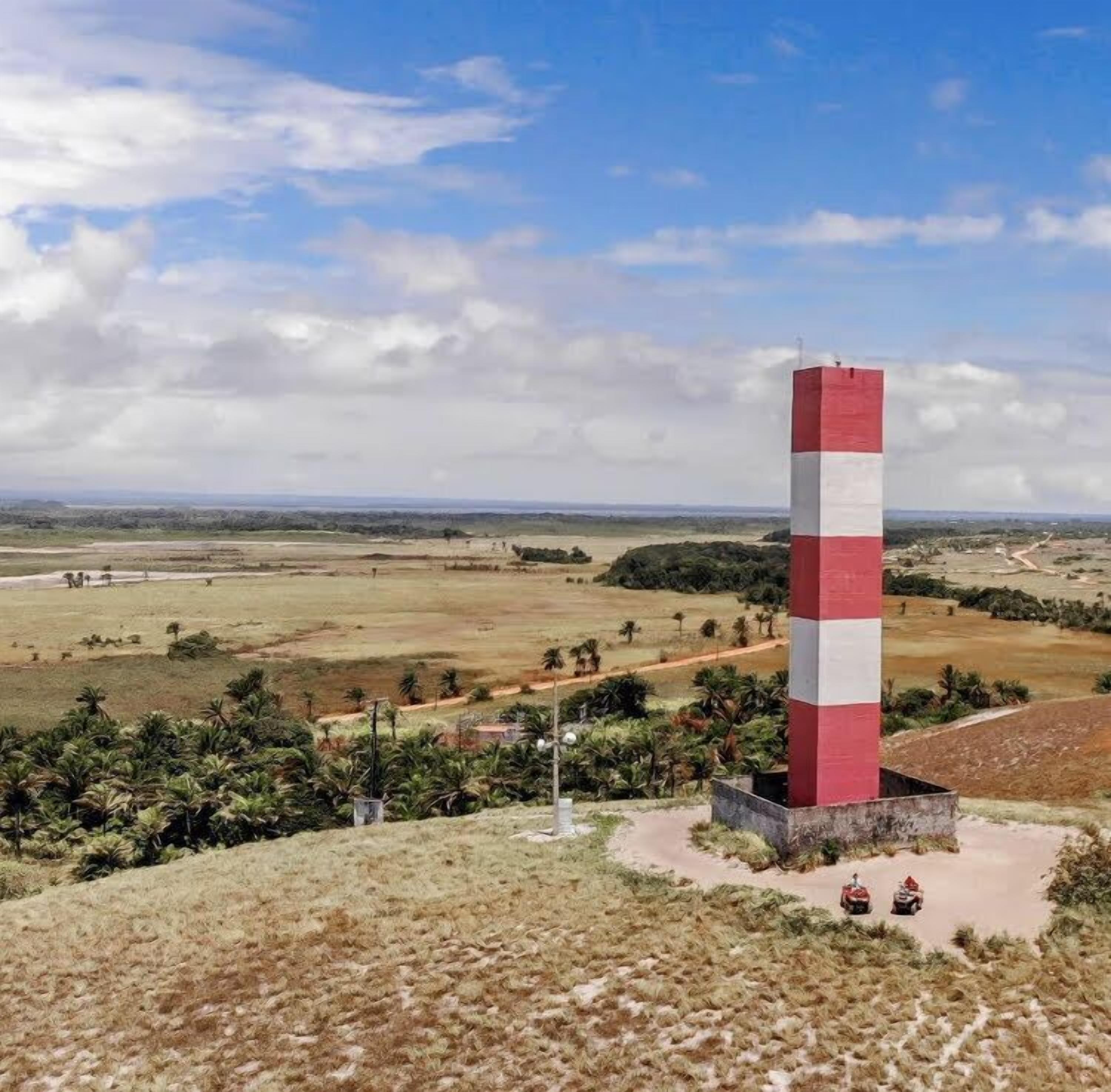 Uma torre alta e esguia com listras vermelhas e brancas alternadas, rodeada por uma paisagem rural com campos e vegetação esparsa.