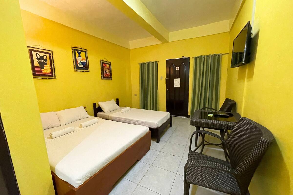 Quarto de hotel com duas camas de solteiro, uma mesinha com cadeiras, televisão e porta com placa.