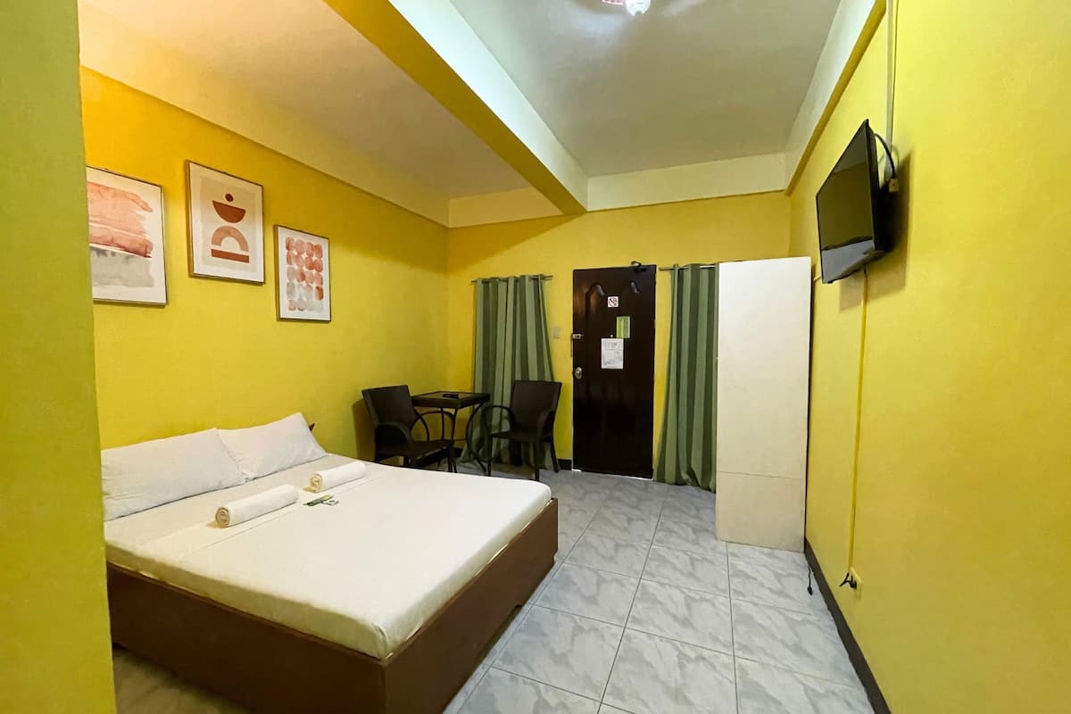 Quarto de hotel com cama, escrivaninha, duas cadeiras, televisão e porta com placa.