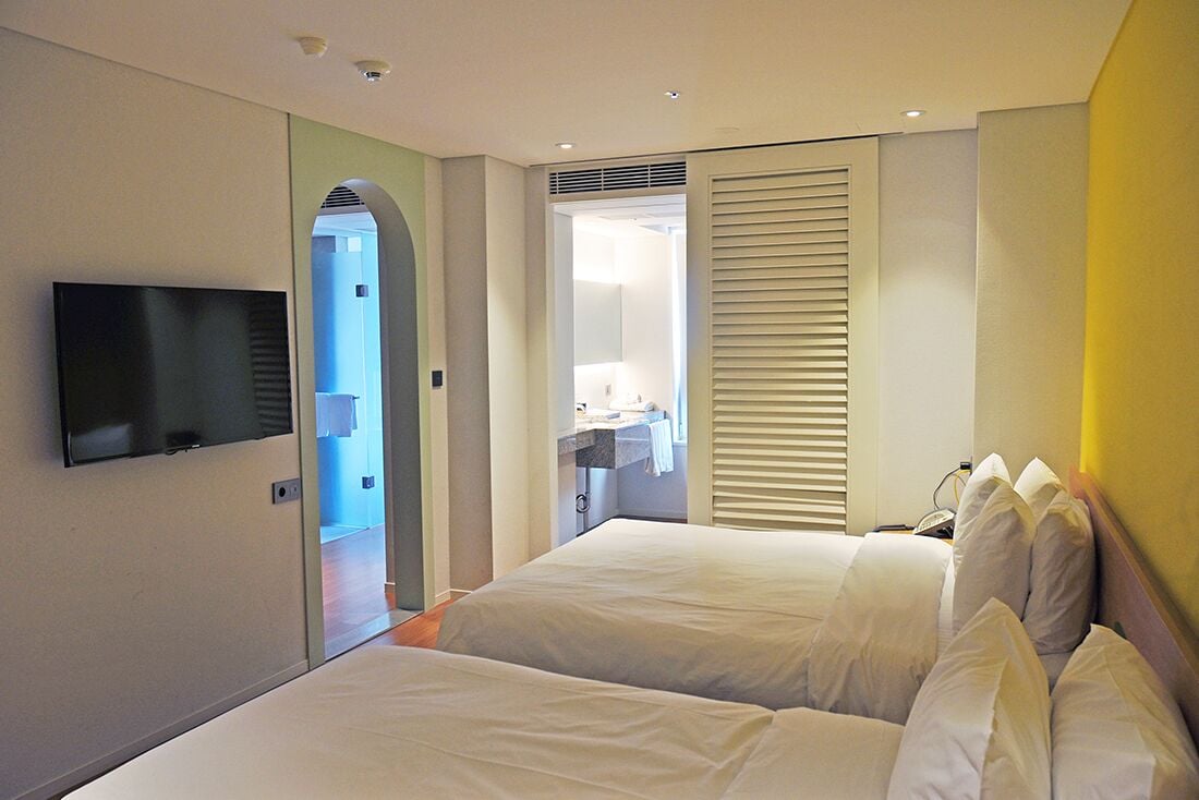 Quarto de hotel com duas camas, televisão presa à parede e banheiro visível por uma porta aberta.