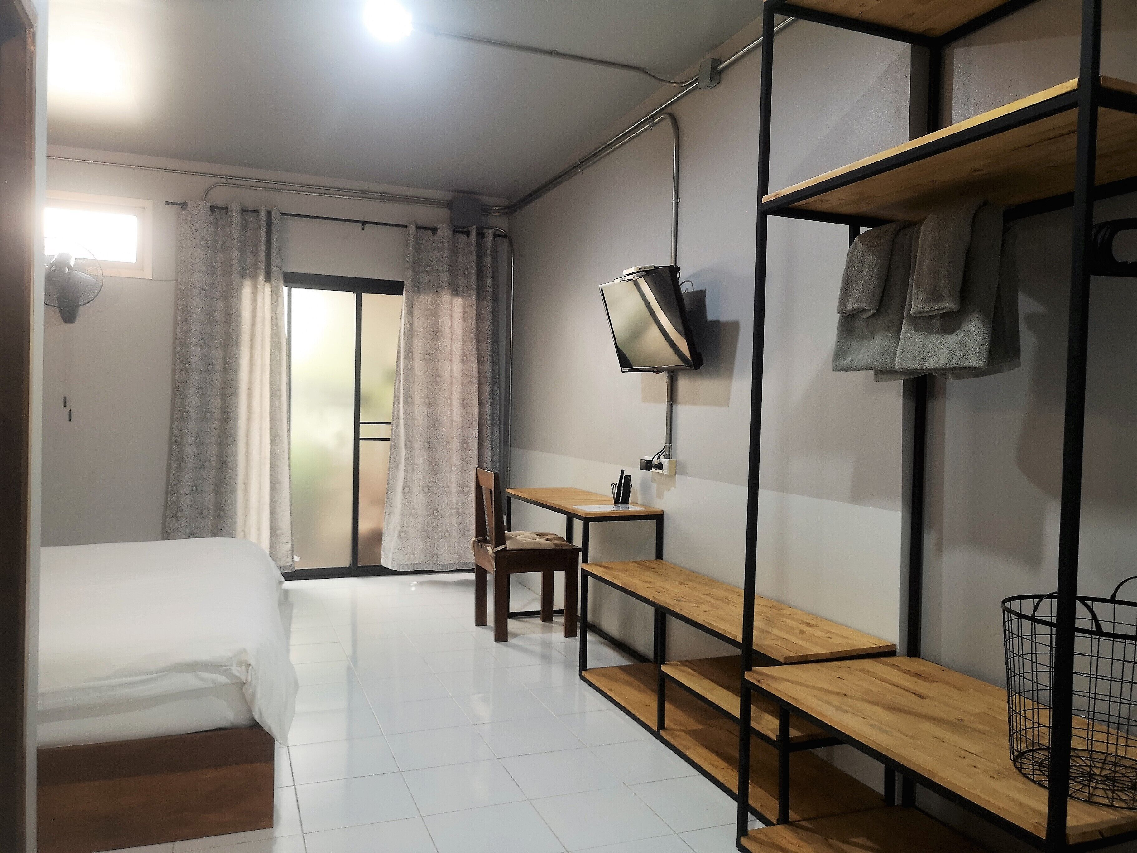 Quarto de hotel com cama, escrivaninha, cadeira, televisão fixada na parede e janela com cortinas.