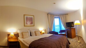 Quarto de hotel com uma cama grande, duas luminárias de cabeceira, uma televisão sobre um móvel de madeira e uma janela com cortinas.