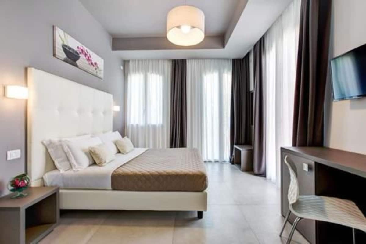 Quarto de hotel moderno com uma cama grande, uma escrivaninha e uma cadeira. Há uma televisão na parede e uma janela com cortinas.