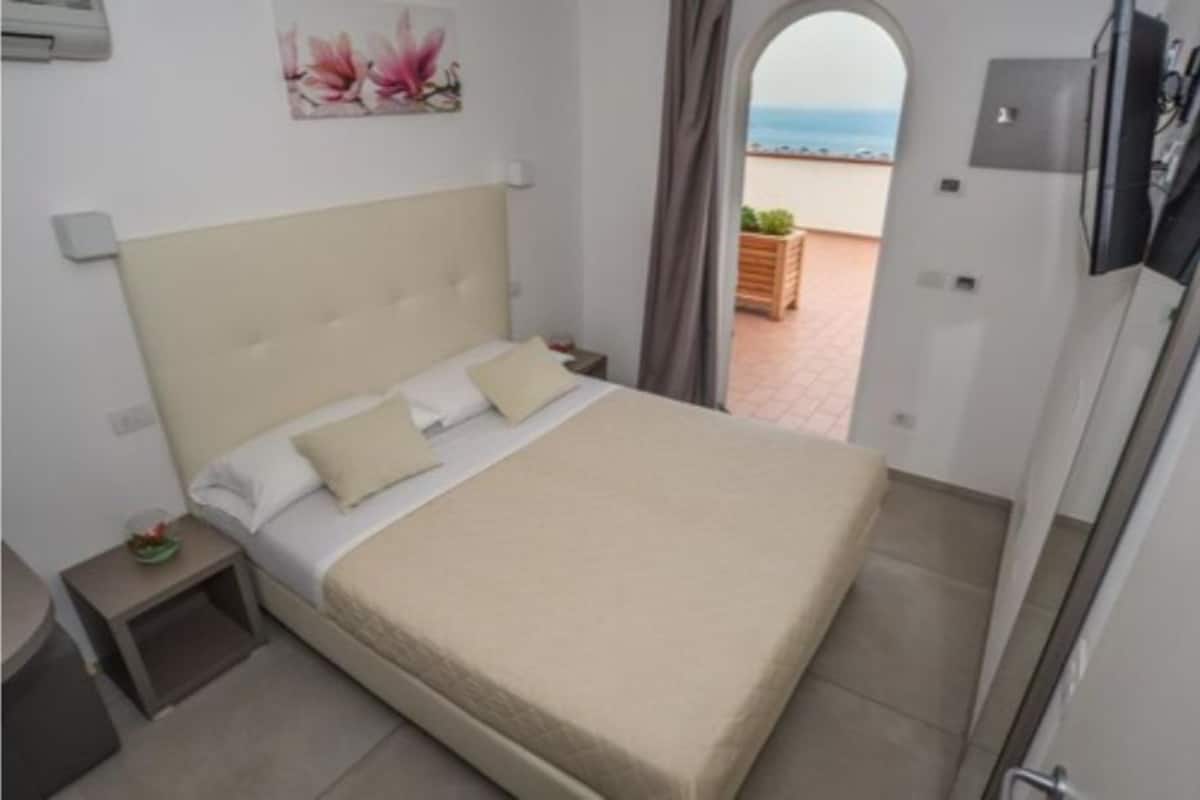 Quarto com cama, uma pequena mesa de cabeceira e vista para a varanda.