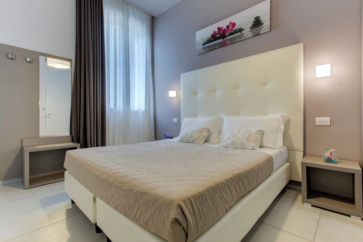 Quarto de hotel moderno com uma cama grande, mesas de cabeceira, um espelho e um quadro na parede.