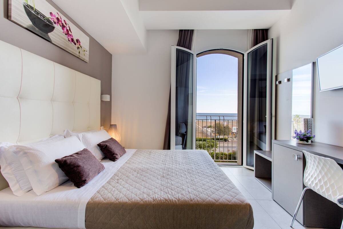 Quarto de hotel com uma cama, uma escrivaninha e vista da cidade através de uma varanda.