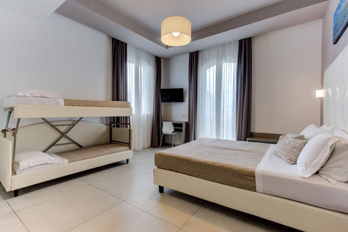 Quarto de hotel com cama de beliche, uma cama de solteiro, escrivaninha com cadeira, televisão de tela plana e uma grande janela com cortinas.