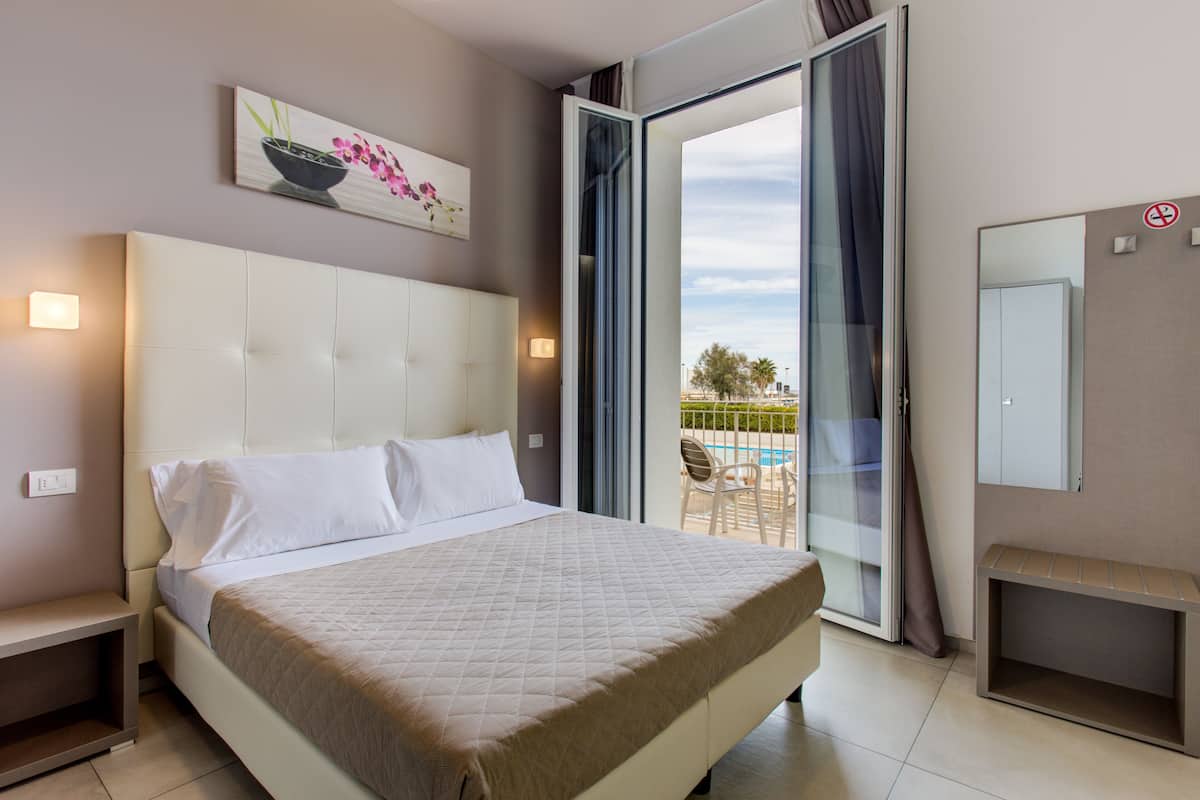 Quarto de hotel moderno com uma cama grande, varanda e vista para a piscina, além de porta de vidro deslizante.