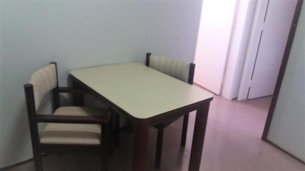 Uma pequena mesa retangular para refeições com quatro cadeiras, cada uma com assento e encosto acolchoados. O quarto tem paredes claras e piso coberto por carpete.
