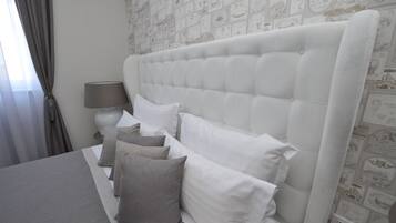 Cama bem arrumada com almofadas brancas e cinzas, cabeceira cinza e abajur em uma mesinha de cabeceira.