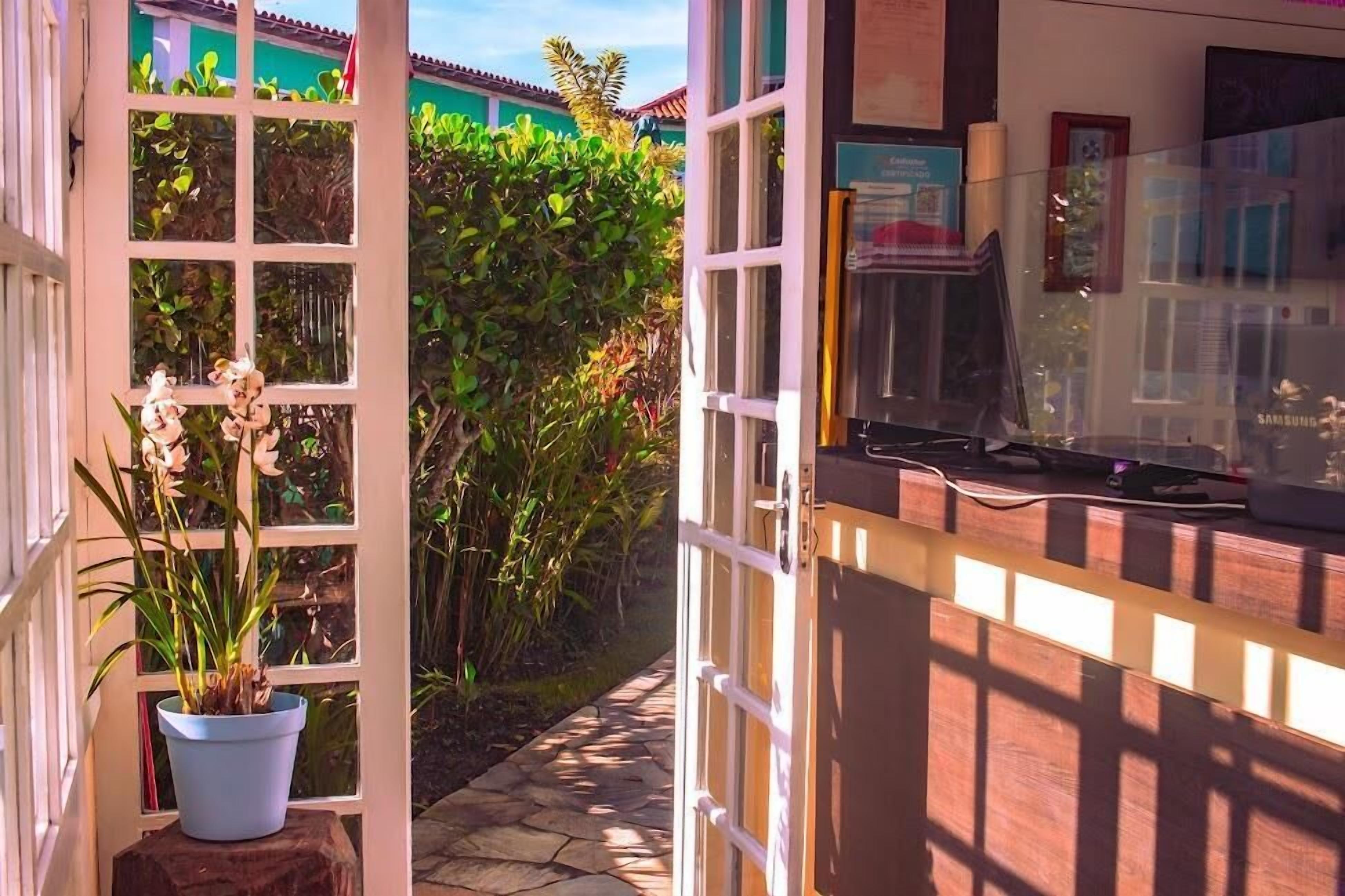 Entrada de hotel com porta de madeira, planta em vaso e uma geladeira Samsung no interior.