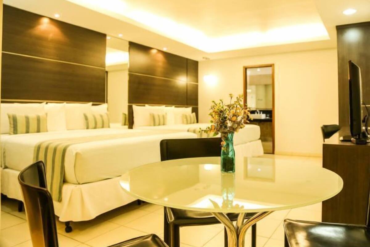 Quarto de hotel com cama, mesa de vidro, um vaso com flores e uma TV.