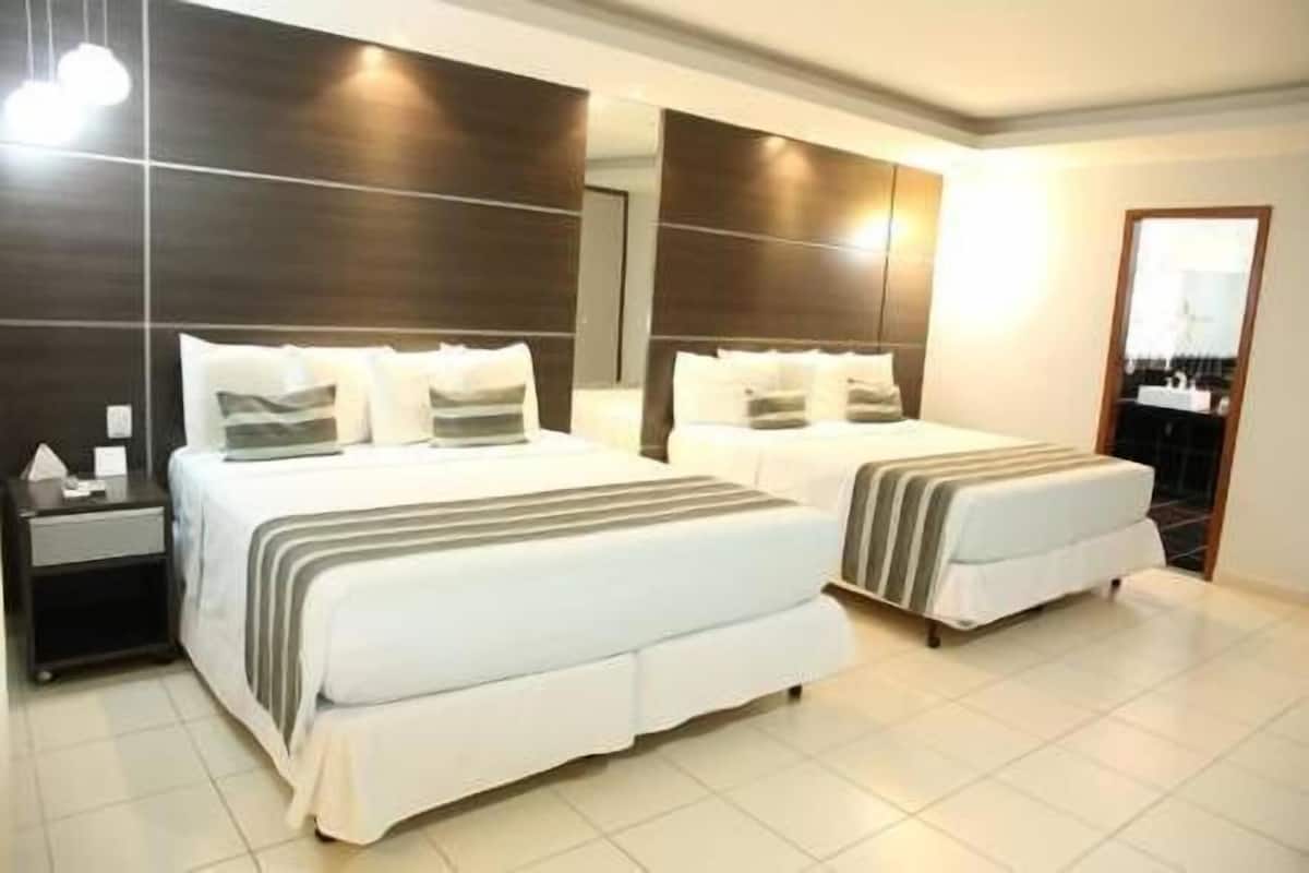 Quarto de hotel com duas camas, uma mesa de cabeceira, um espelho e um banheiro visível por uma porta aberta.