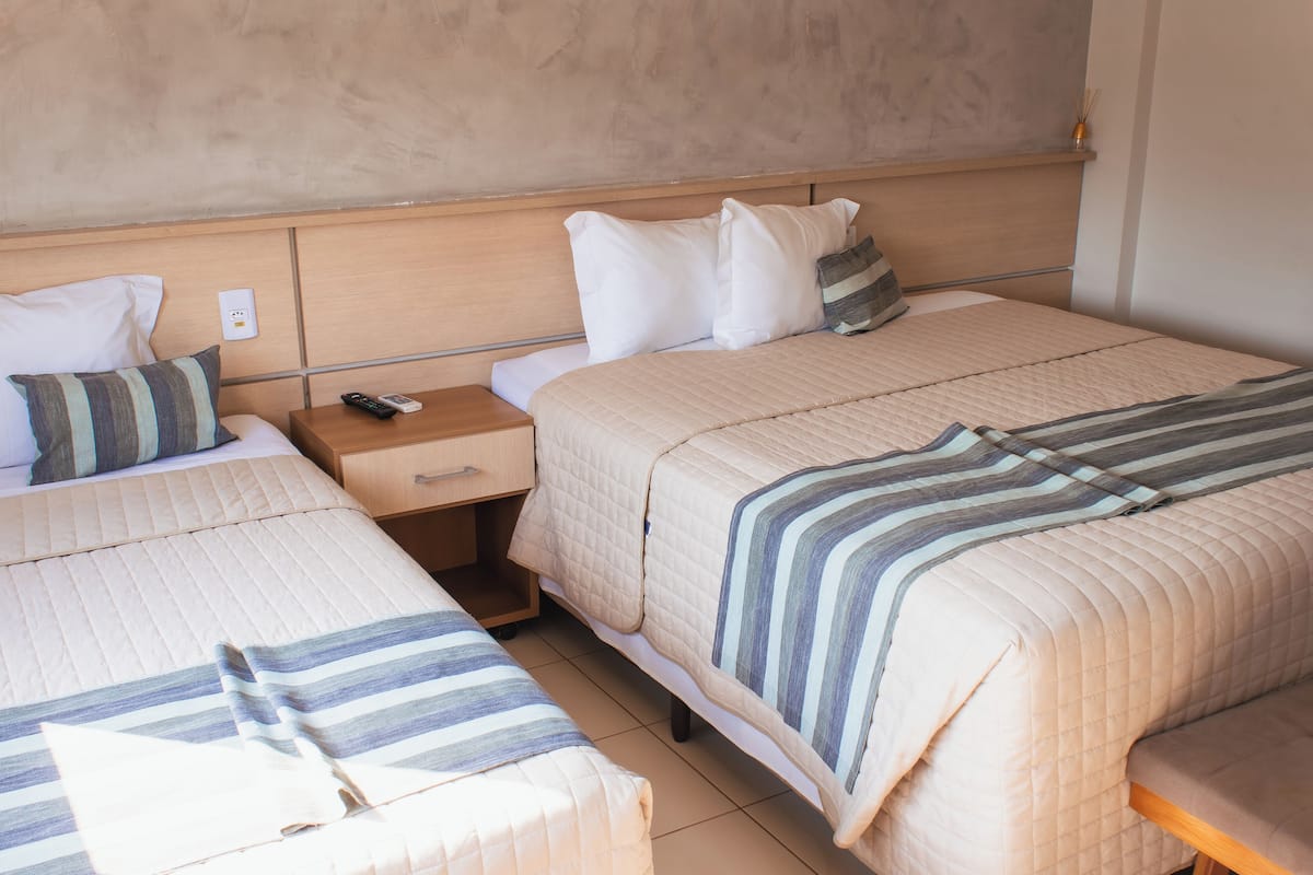 Quarto de hotel com duas camas, cada uma com um cobertor listrado diferente, uma cabeceira de madeira e uma mesinha de cabeceira com um controle remoto.