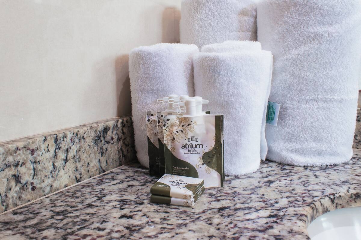 Casa de banho com toalhas brancas empilhadas na parede, um frasco de produtos de banho do Hotel Atrium e bancada de mármore.