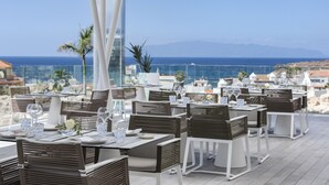 Restaurante com área externa e mesas ao ar livre, com vista para o mar, com mesas brancas e cadeiras marrons, e uma vista desobstruída da costa e montanhas ao fundo.