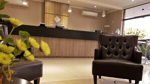Uma área de recepção de hotel com um balcão de madeira, uma poltrona de couro preto e um vaso com flores amarelas.