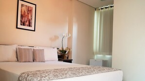 Quarto de hotel com uma cama grande, um quadro na parede, uma mesa de cabeceira com um vaso de flores e uma janela com cortinas.