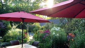 Um pátio externo com mesa e cadeiras de madeira, cercado por vegetação exuberante e plantas floridas.