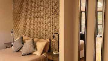 Quarto com papel de parede estampado, uma cama com almofadas, mesa de cabeceira com abajur e vista pela janela.