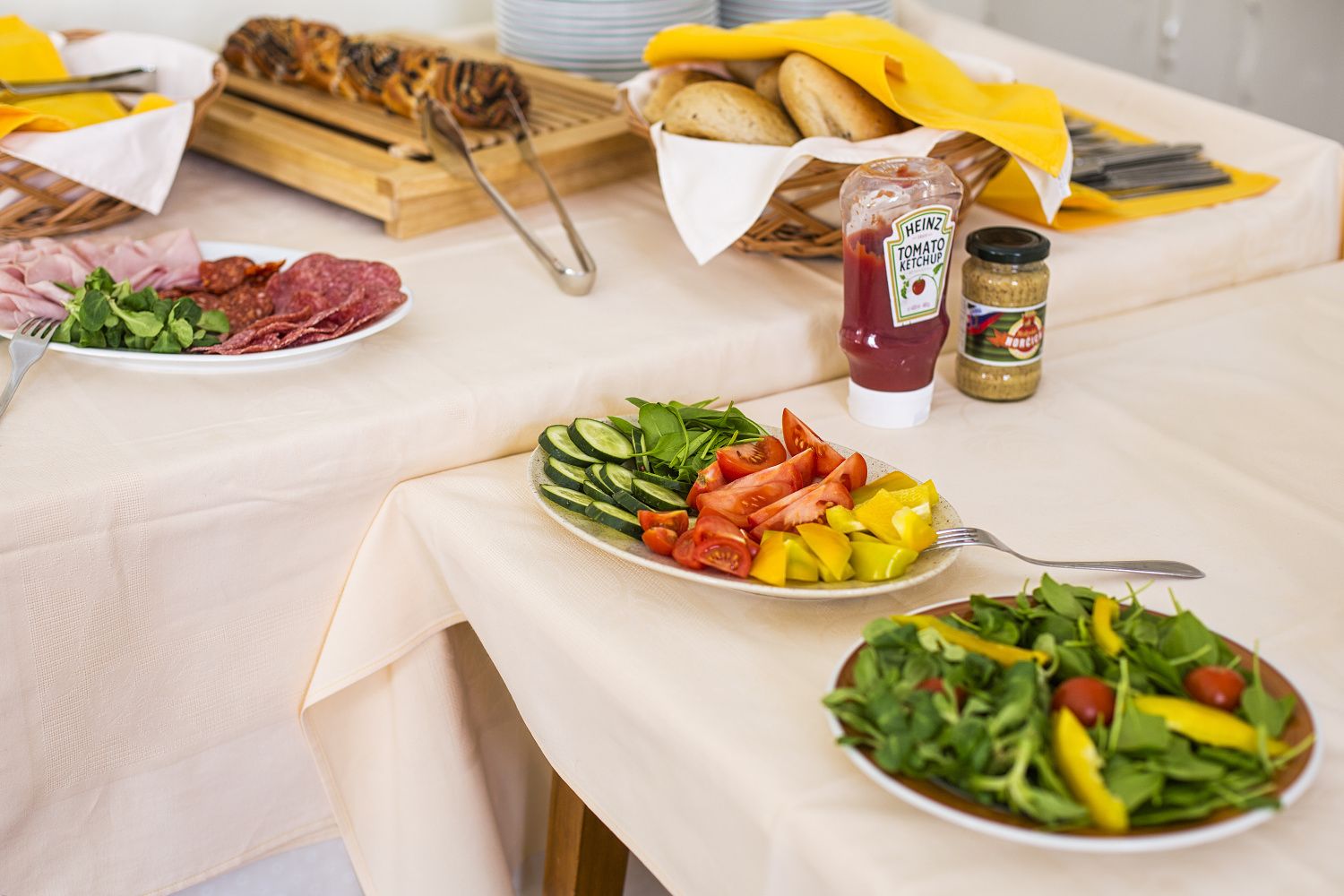 Uma mesa com diversos alimentos, incluindo pepinos fatiados, tomates, pimentões amarelos e uma salada com folhas verdes e tomates cereja. Há também acompanhamentos como ketchup Heinz e um pote de mostarda.