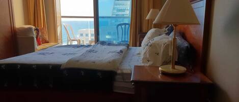 Quarto de hotel com uma cama, uma mesa de cabeceira com abajur, uma cadeira e uma janela com vista para um edifício e o mar.