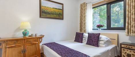 Quarto com cama, cómoda de madeira, candeeiro, janela com cortinas e um quadro na parede.