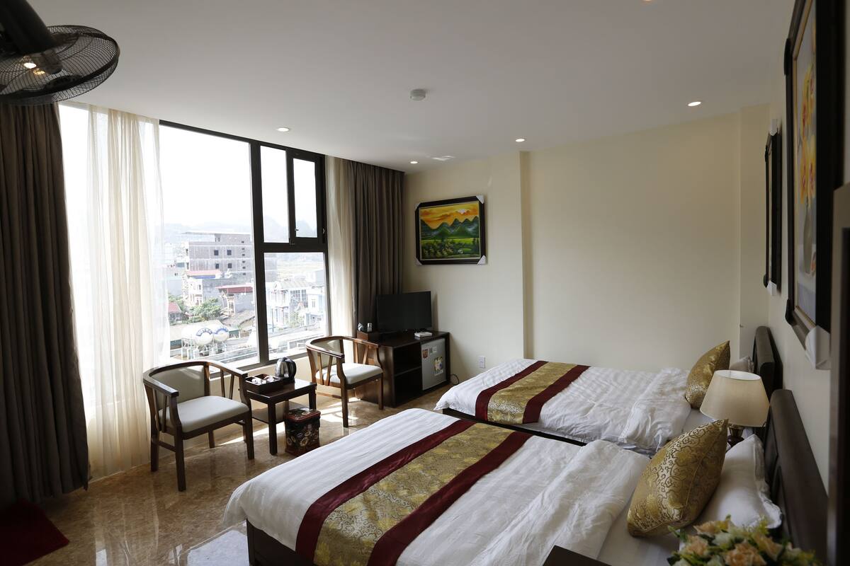 Quarto de hotel com duas camas, uma escrivaninha, uma cadeira, televisão e uma janela grande com vista para a cidade.