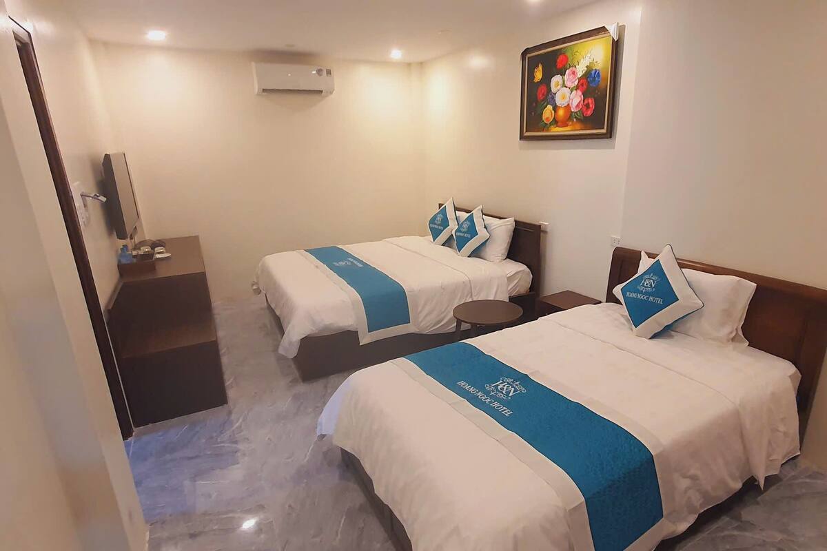 Quarto de hotel com duas camas, cada uma com colcha e almofadas azuis, uma pequena mesa de cabeceira, televisão presa à parede e um quadro sobre as camas.