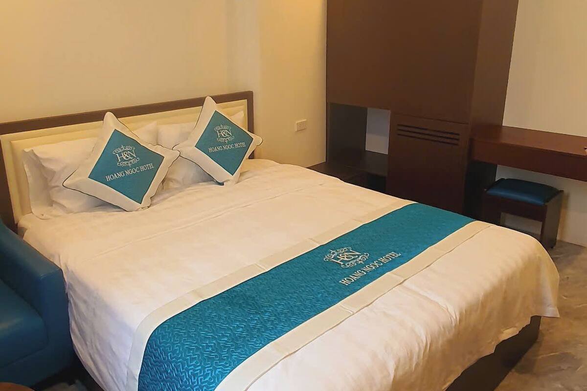 Quarto de hotel com cama arrumada, uma cadeira azul, uma mesinha e um quadro na parede.