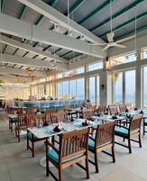 Um restaurante com vista para o mar, cadeiras e mesas de madeira, ventiladores de teto e luminárias pendentes.