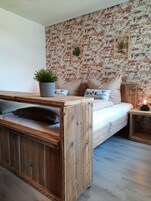 Um quarto aconchegante com uma estrutura de cama de madeira rústica, uma pequena mesa de cabeceira com abajur e uma parede com padrão de tijolos e folhas emolduradas.