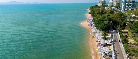 Uma praia com uma fileira de guarda-sóis e cadeiras de praia, uma estrada ao lado da orla e edifícios altos ao fundo.