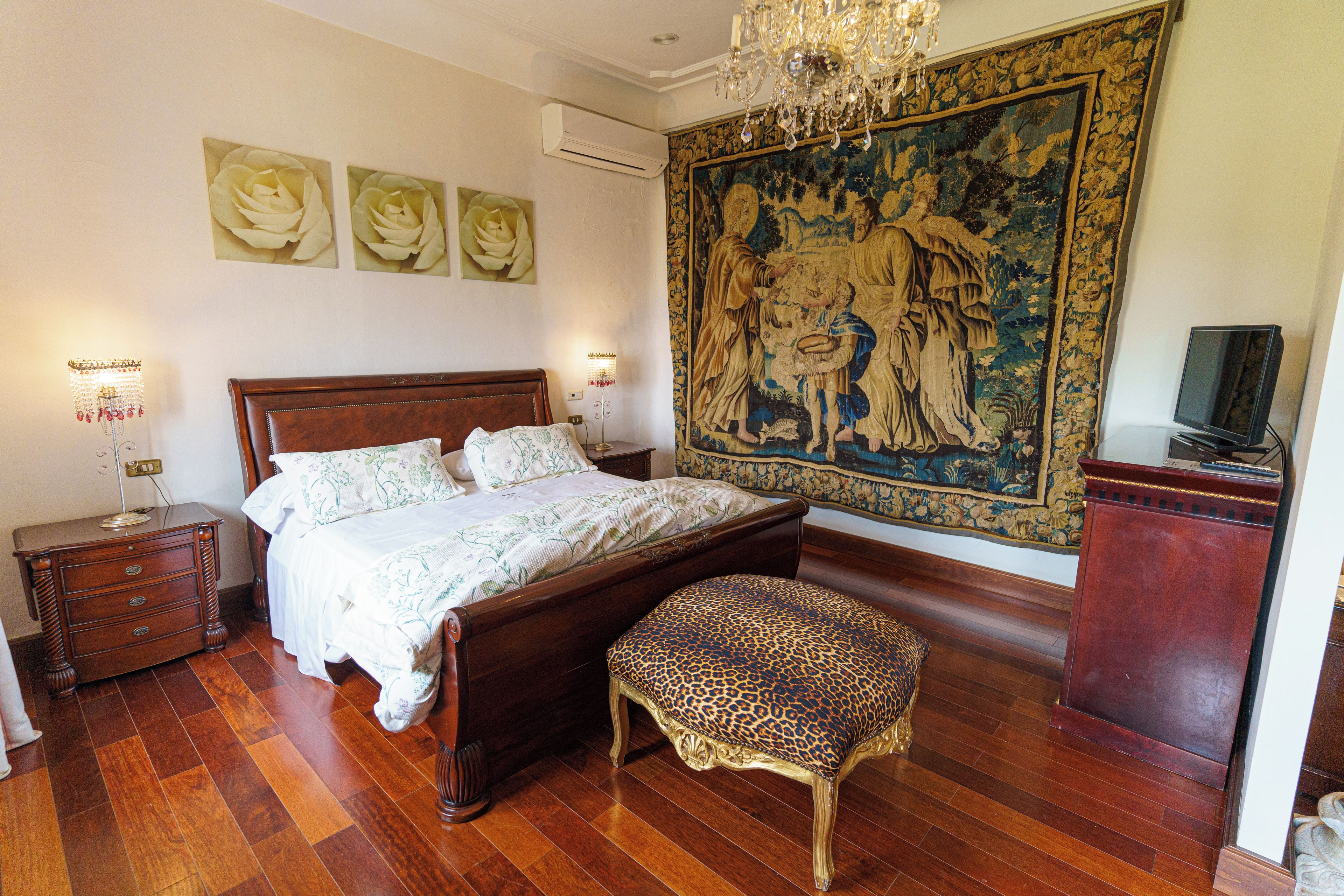 Quarto com cama grande, mesa de cabeceira, abajur, banquinho e tapeçaria na parede.