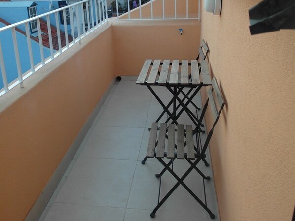 Uma varanda com mesa e cadeiras de madeira, guarda-corpo branco e vista para prédios ao fundo.