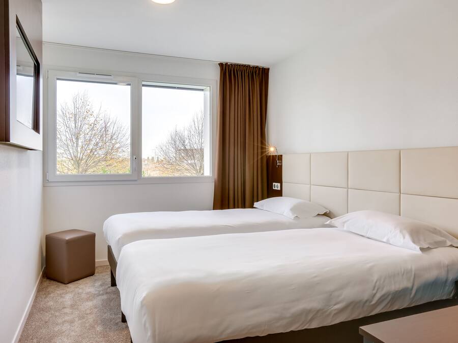 Quarto de hotel com duas camas, janela com cortinas, mesa de cabeceira e um banquinho pequeno.
