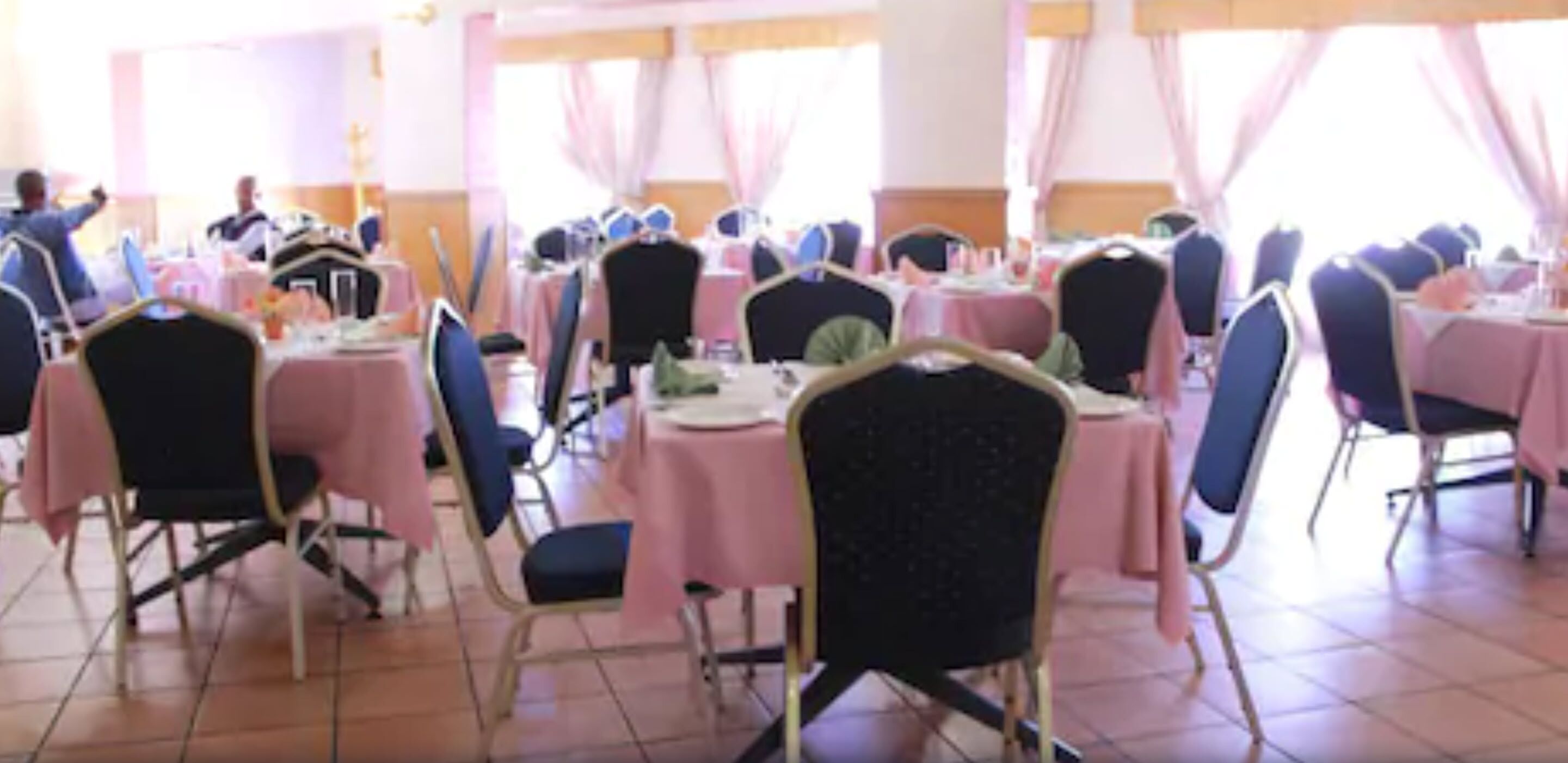 Salão de banquetes com mesas redondas cobertas por toalhas cor-de-rosa e cadeiras azuis.