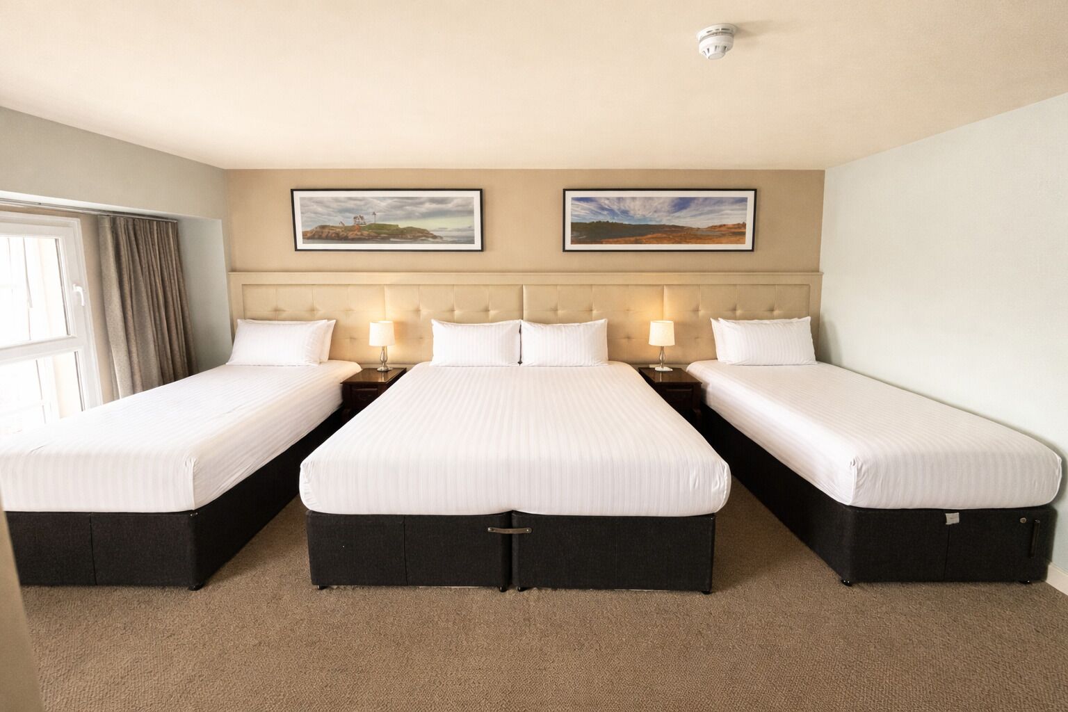 Quarto de hotel com três camas de solteiro, cabeceira com design estofado, duas pinturas emolduradas de paisagens e detector de fumaça no teto.