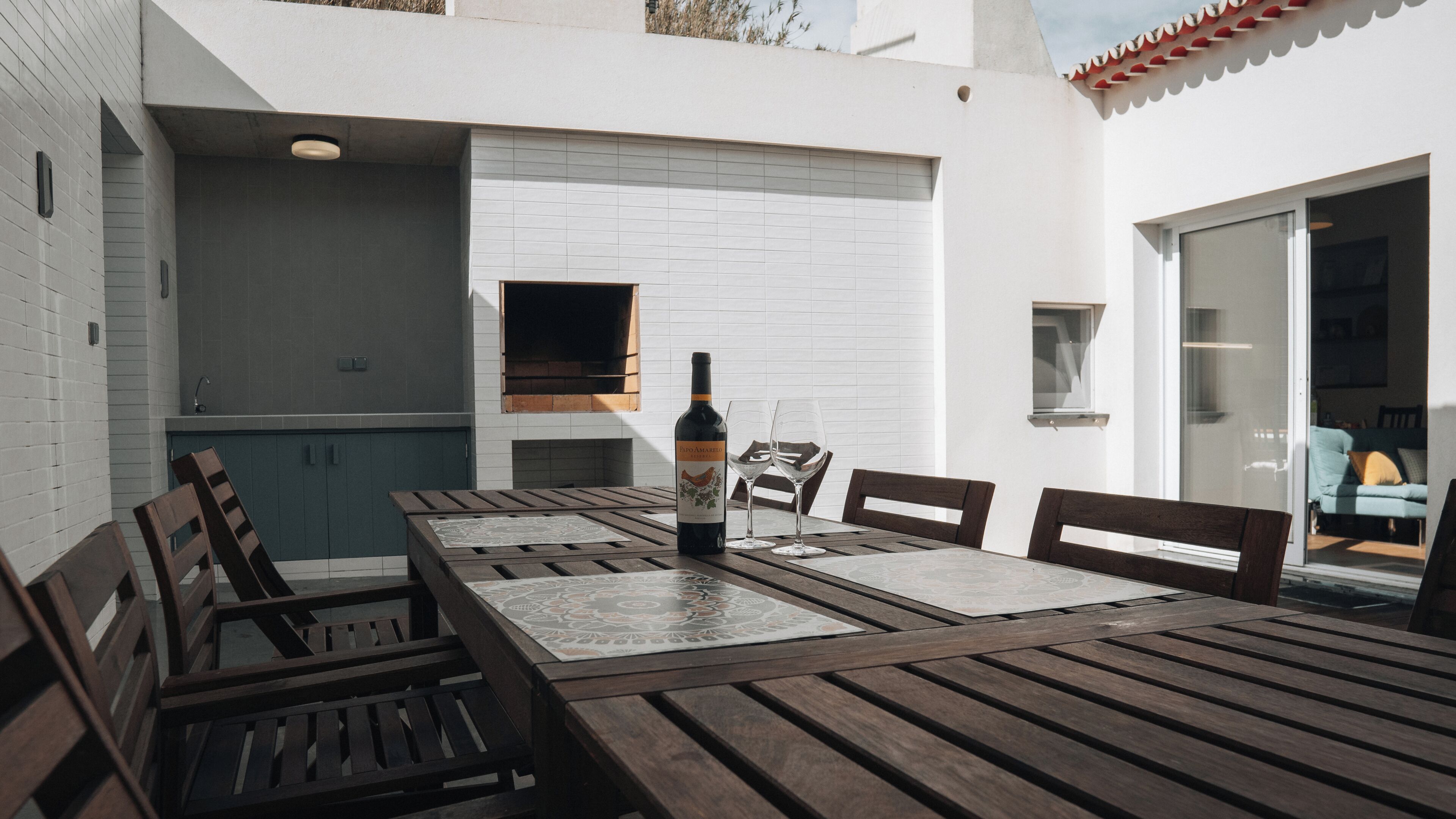 Uma área de refeições ao ar livre com mesa, cadeiras, uma garrafa de vinho e vista para uma casa.