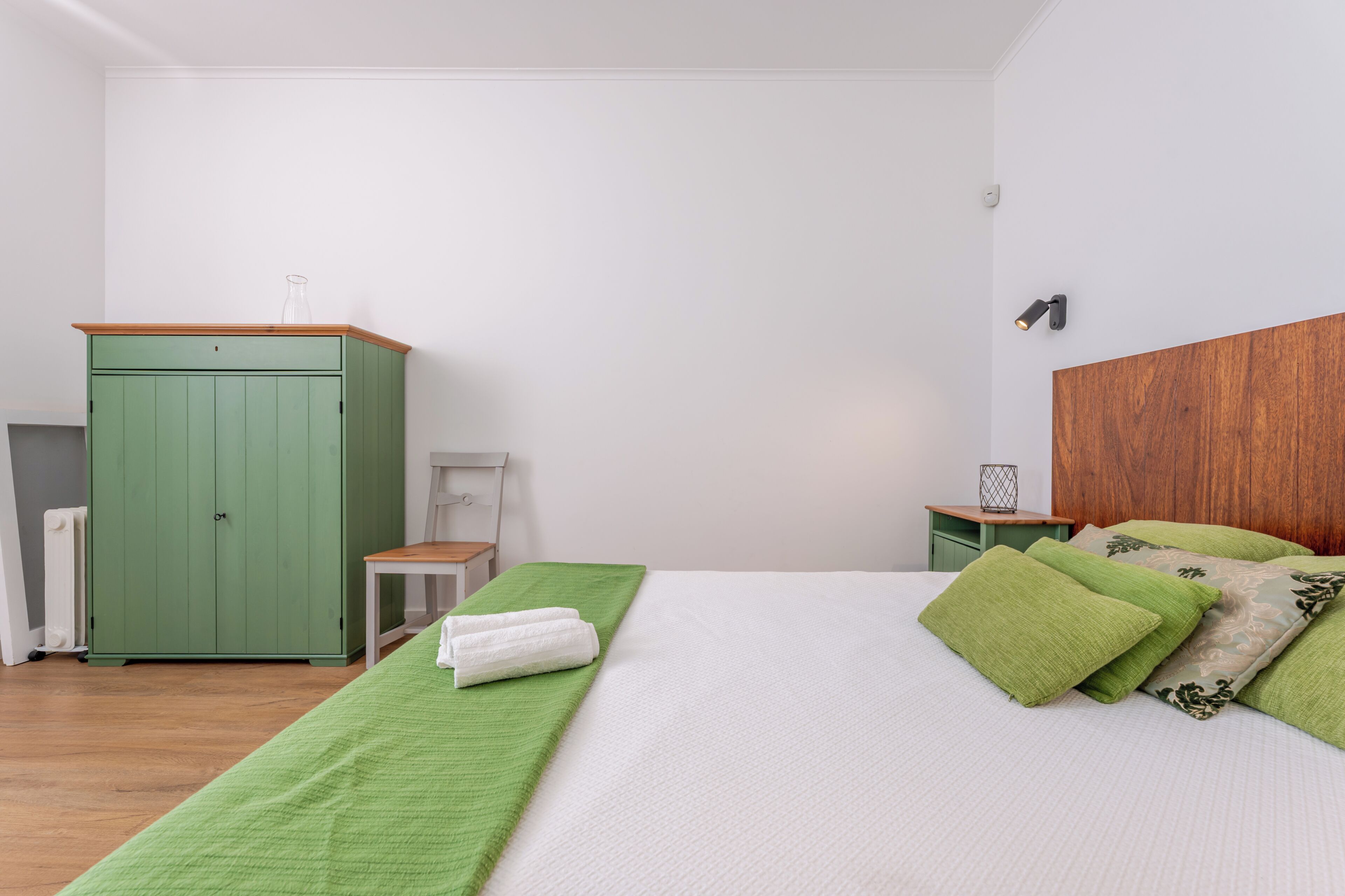 Quarto com cama, colcha verde, almofadas brancas, guarda-roupa verde, cabeceira de madeira e mesa de cabeceira com abajur.