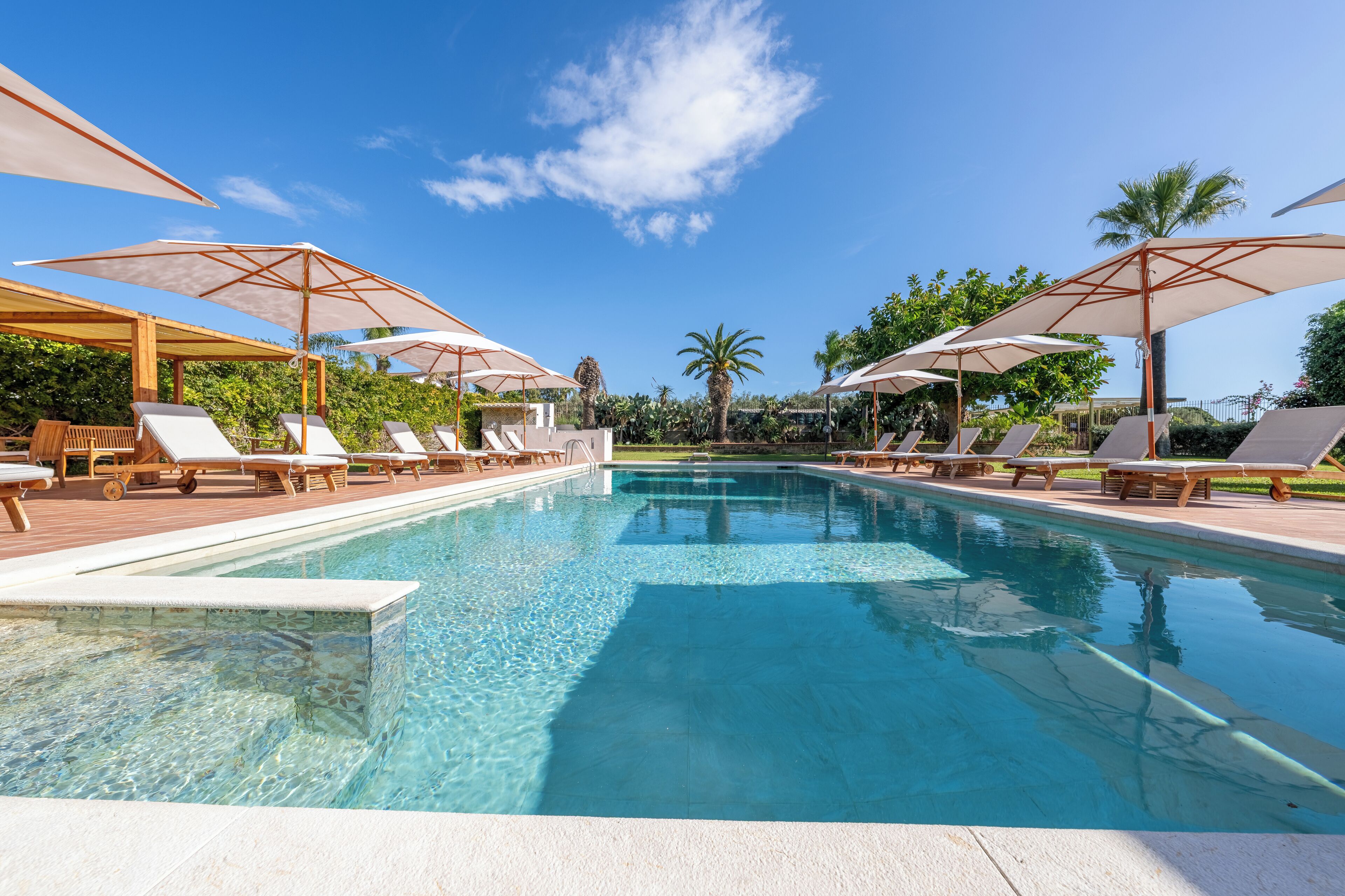 Piscina límpida com espreguiçadeiras e guarda-sóis, cercada por vegetação e palmeiras.