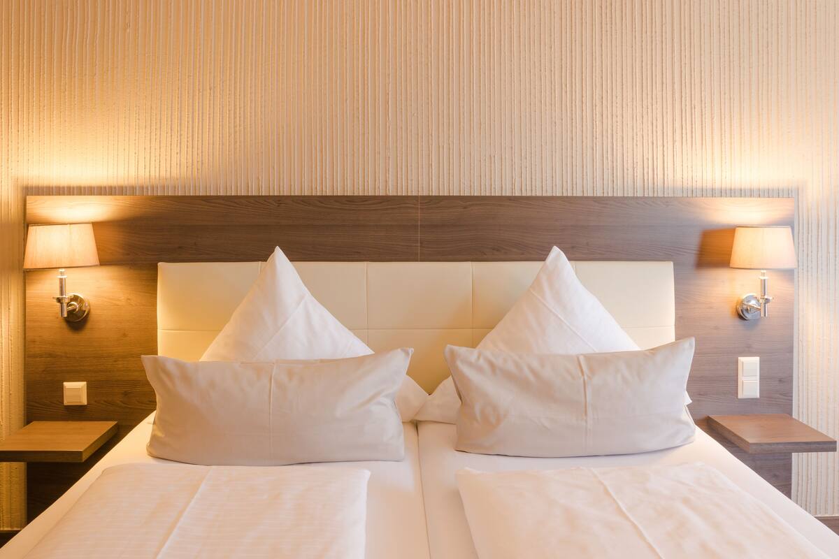 Uma cama de casal com duas almofadas brancas, uma cabeceira de madeira e duas luminárias de cabeceira.