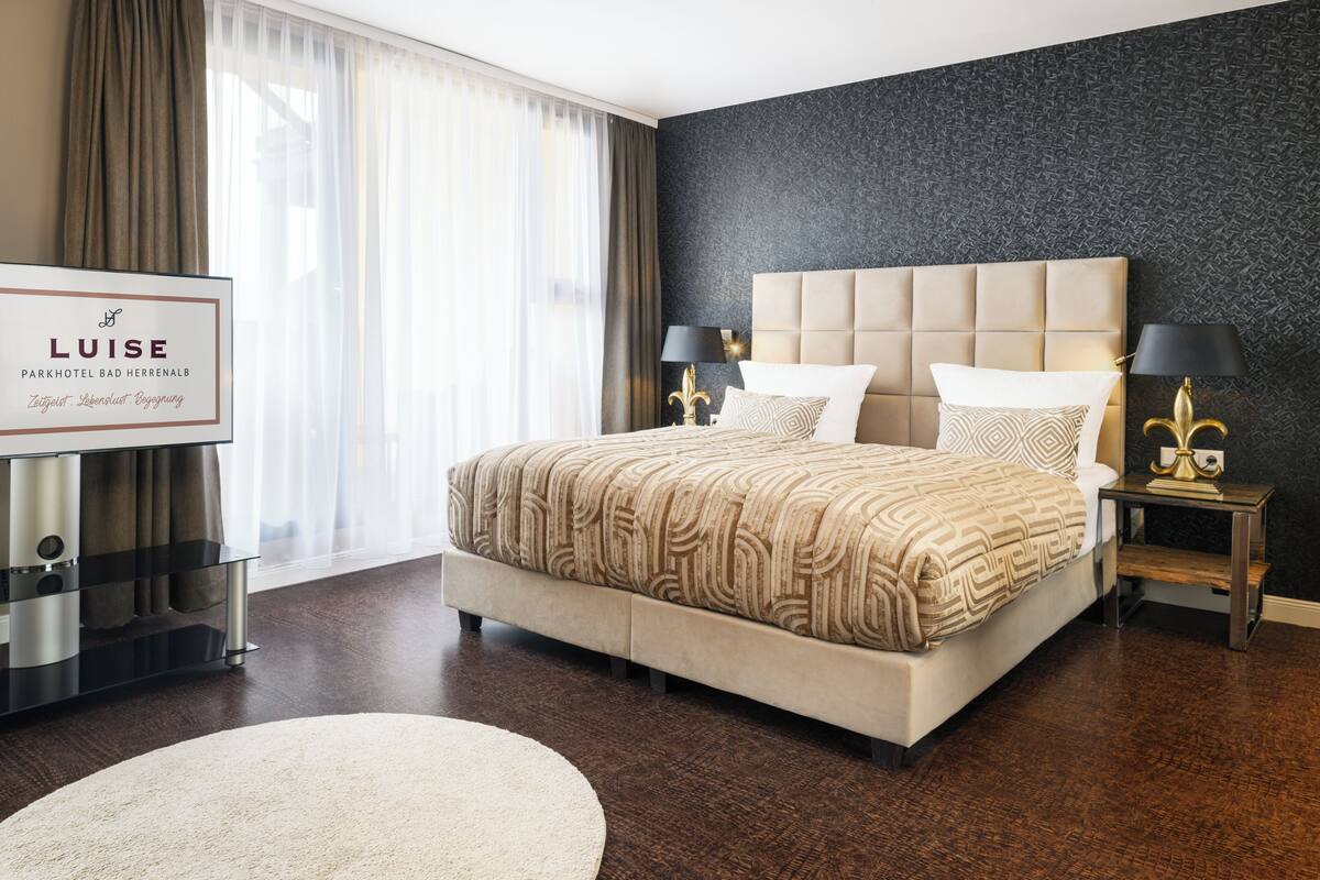 Quarto de hotel com uma cama grande, piso de madeira escura, parede texturizada em tons escuros e uma janela ampla com cortinas.