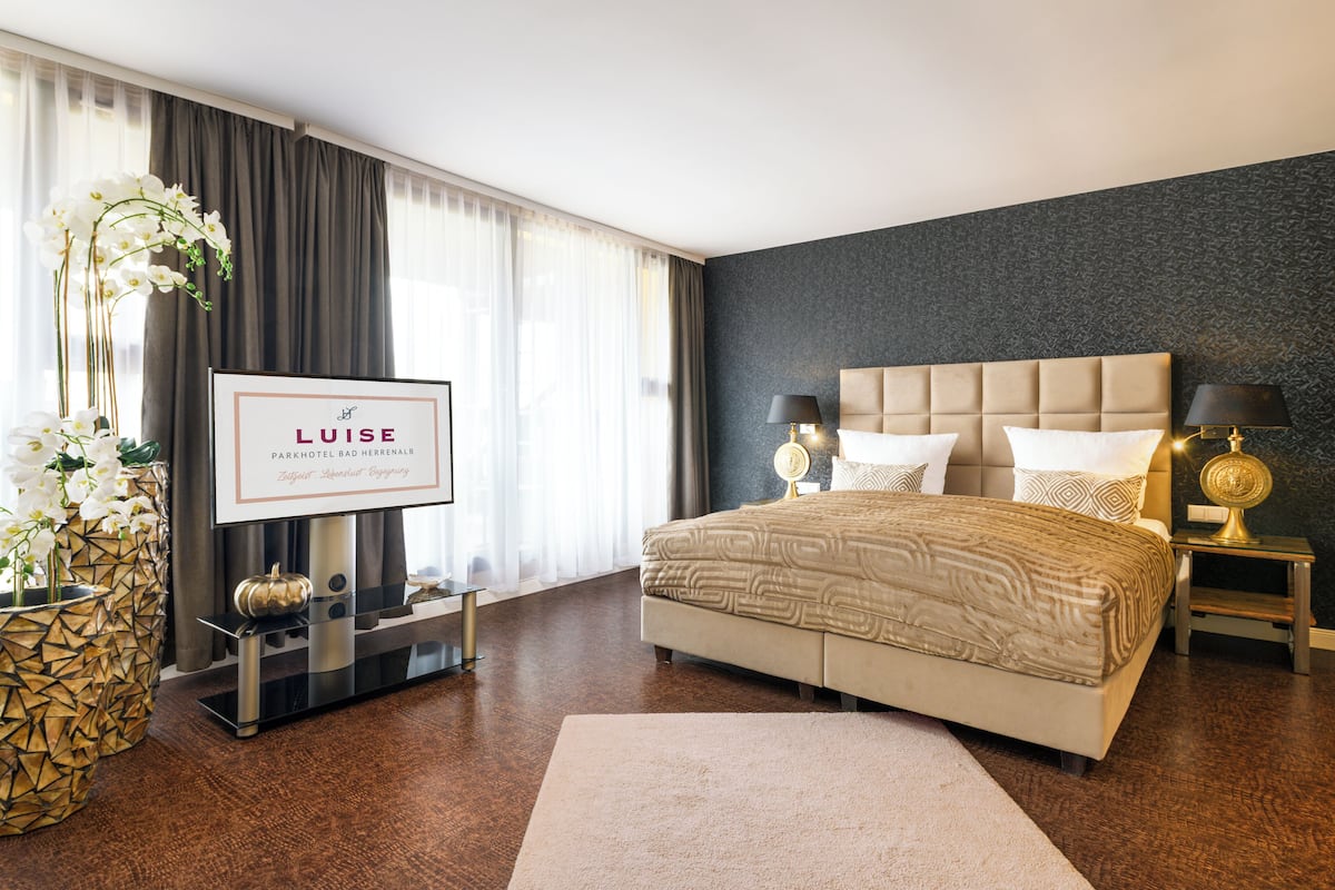 Quarto de hotel moderno com uma cama grande, uma televisão, um vaso com flores e uma parede escura.