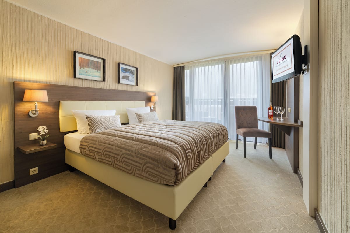 Quarto de hotel com uma cama grande, mesas de cabeceira, uma cadeira, televisão e janela com cortinas.