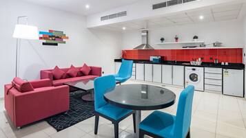 Uma sala de estar moderna com um sofá vermelho, cadeiras azuis e uma mesa de vidro. Uma cozinha com azulejos vermelhos e armários brancos é visível ao fundo.