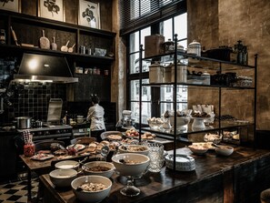 Um chef prepara comida em uma cozinha com uma janela grande, prateleiras com utensílios de cozinha e diversos pratos sobre o balcão.