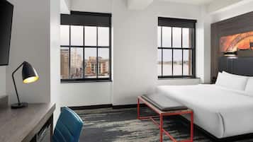 Quarto de hotel com cama, escrivaninha, cadeira, luminária de mesa e duas janelas com vista para a cidade.