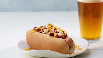 Um cachorrinho com chili e queijo no pão, servido em um prato branco, com um copo de cerveja ao fundo.